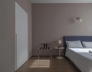 Quarto com cama, cadeira, mesa pequena, luminária na parede e guarda-roupa.