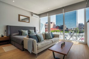 Quarto moderno com cama grande, sofá, mesa de centro de madeira e vista para a cidade através de grandes janelas.