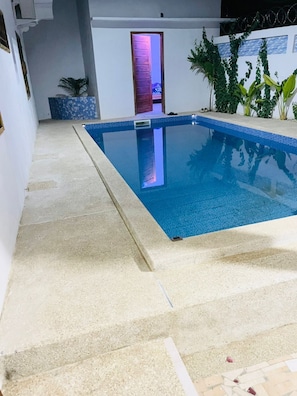Piscina retangular com deck ladrilhado, rodeada por paredes brancas e plantas em vasos.
