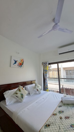 Quarto de hotel com cama, ventoinha no teto, ar condicionado, janela com cortinas e um quadro pendurado na parede.