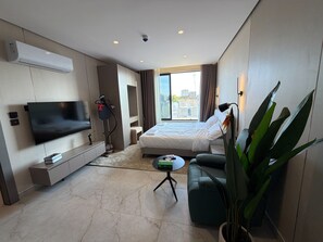 Quarto de hotel moderno com televisão de tela plana, uma cama, sofá, mesa de centro e uma planta grande.