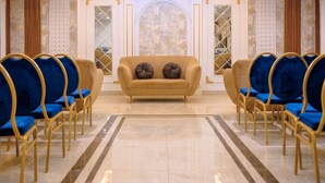 Um saguão de hotel com um sofá central, cadeiras azuis, um candelabro e uma parede estampada.