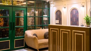 Um hall com balcão de recepção, porta de vidro com moldura verde, sofá bege e um relógio na parede.