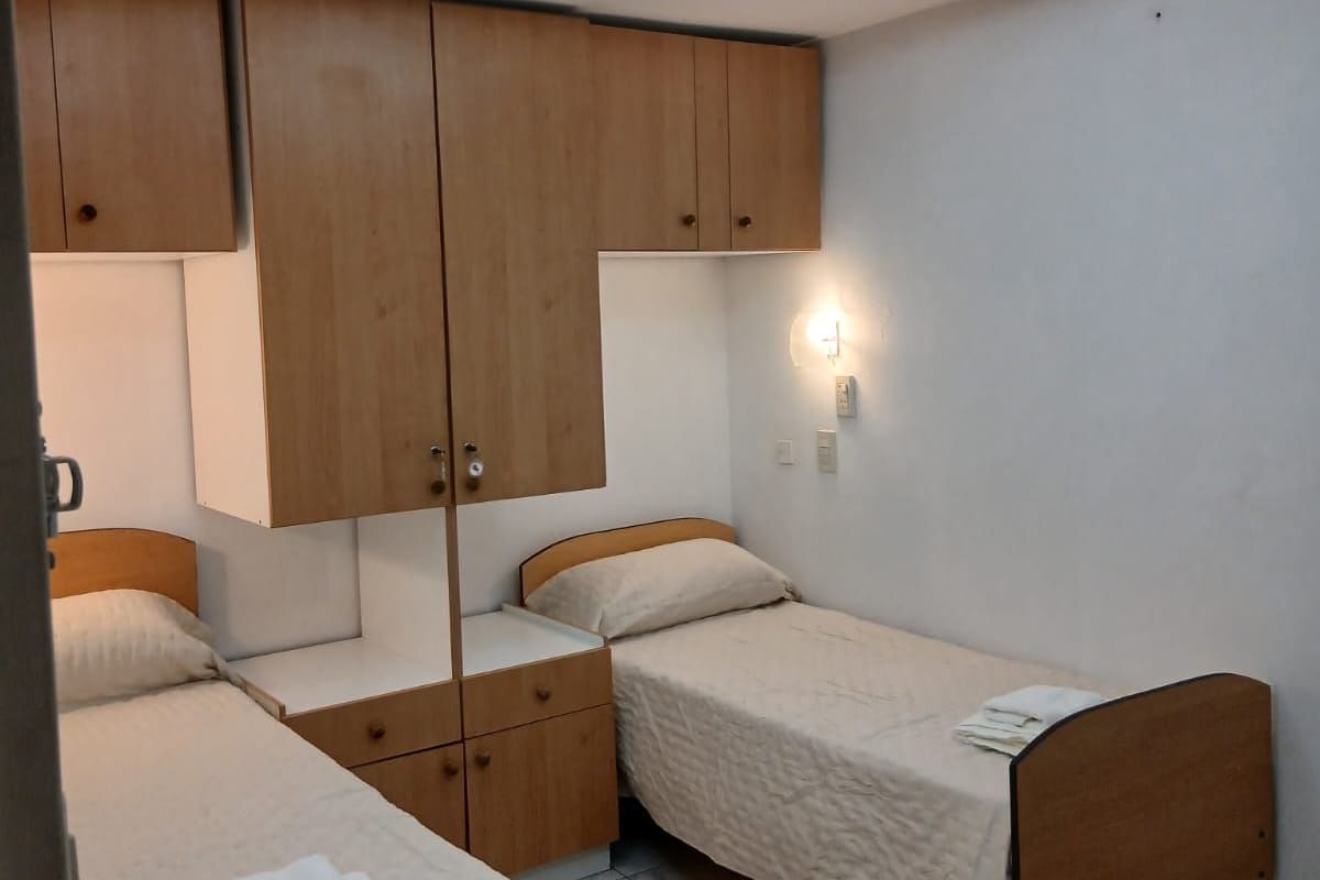 Quarto compacto com duas camas de solteiro, armários de madeira, uma mesinha e luminária no teto.