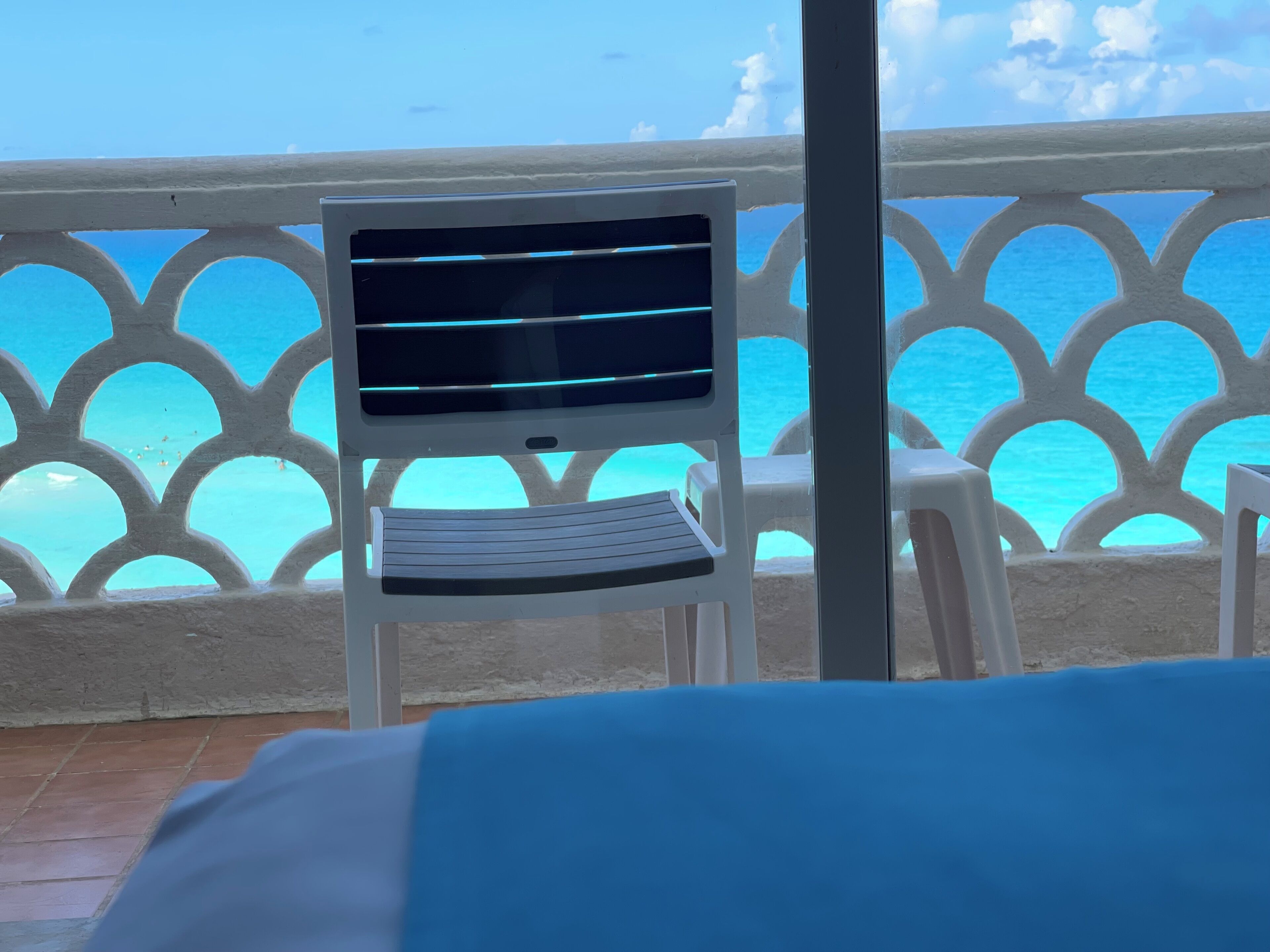 Uma cadeira de plástico branca com encosto de ripas e uma mesa pequena, posicionada numa varanda com vista para o oceano.