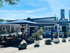 Uma área de estar ao ar livre com mesas e cadeiras, um café e bar de vidro, e uma loja Butik delicatessen.