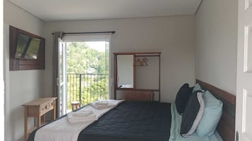 Quarto com cama, mesa de cabeceira, espelho e janela com vista para o verde.