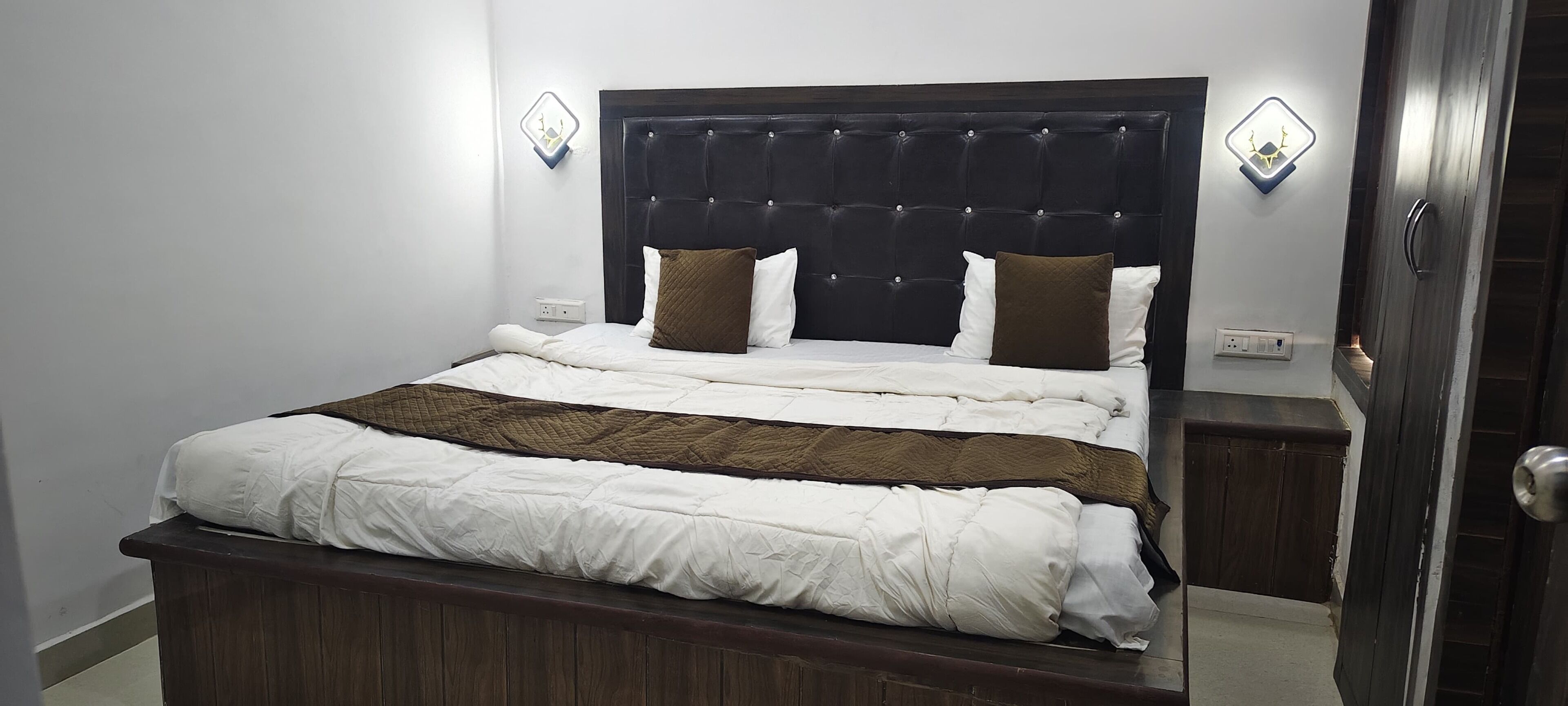 Cama bem arrumada com cabeceira escura, duas mesas de cabeceira e luminárias fixadas na parede.