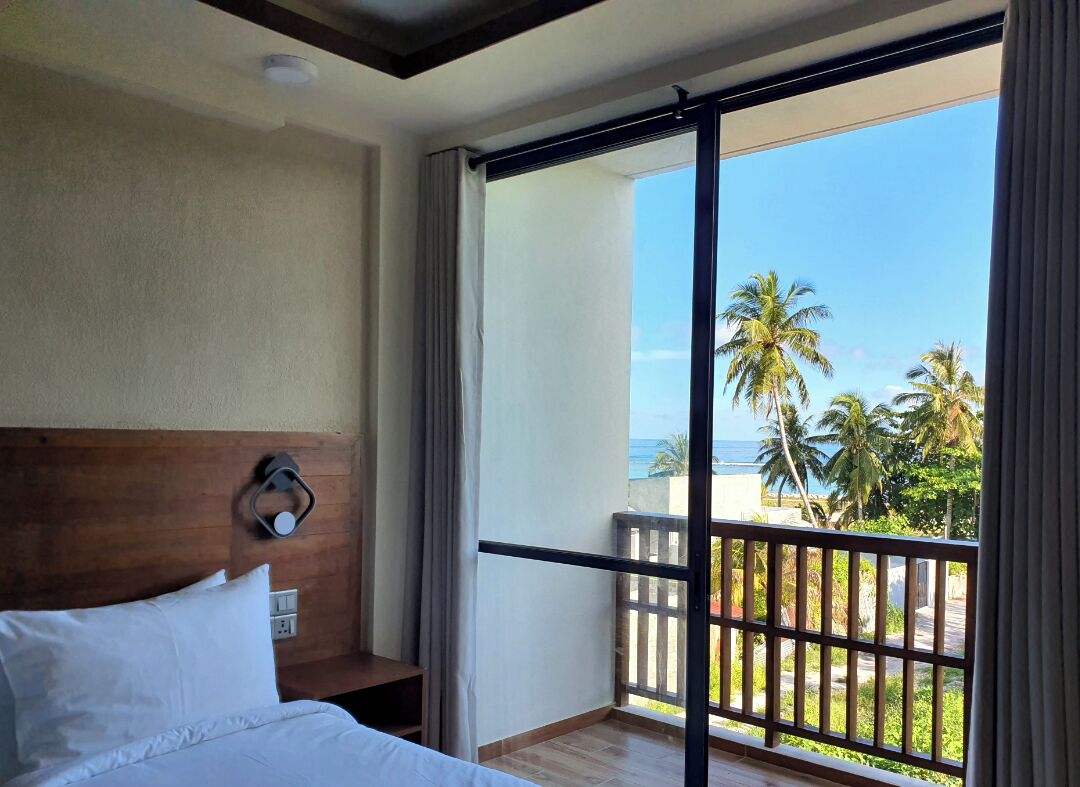 Quarto de hotel com cama, cabeceira de madeira, janela com vista para palmeiras e oceano, e varanda com guarda-corpo.