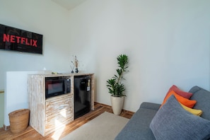 Sala de estar com micro-ondas embutido, planta em vaso, sofá cinza com almofadas coloridas e TV na parede exibindo 'NETFLIX'.
