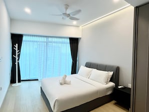 Quarto de hotel moderno com uma cama grande, mesa de cabeceira, ventilador de teto e vista para edifícios altos através de cortinas translúcidas.