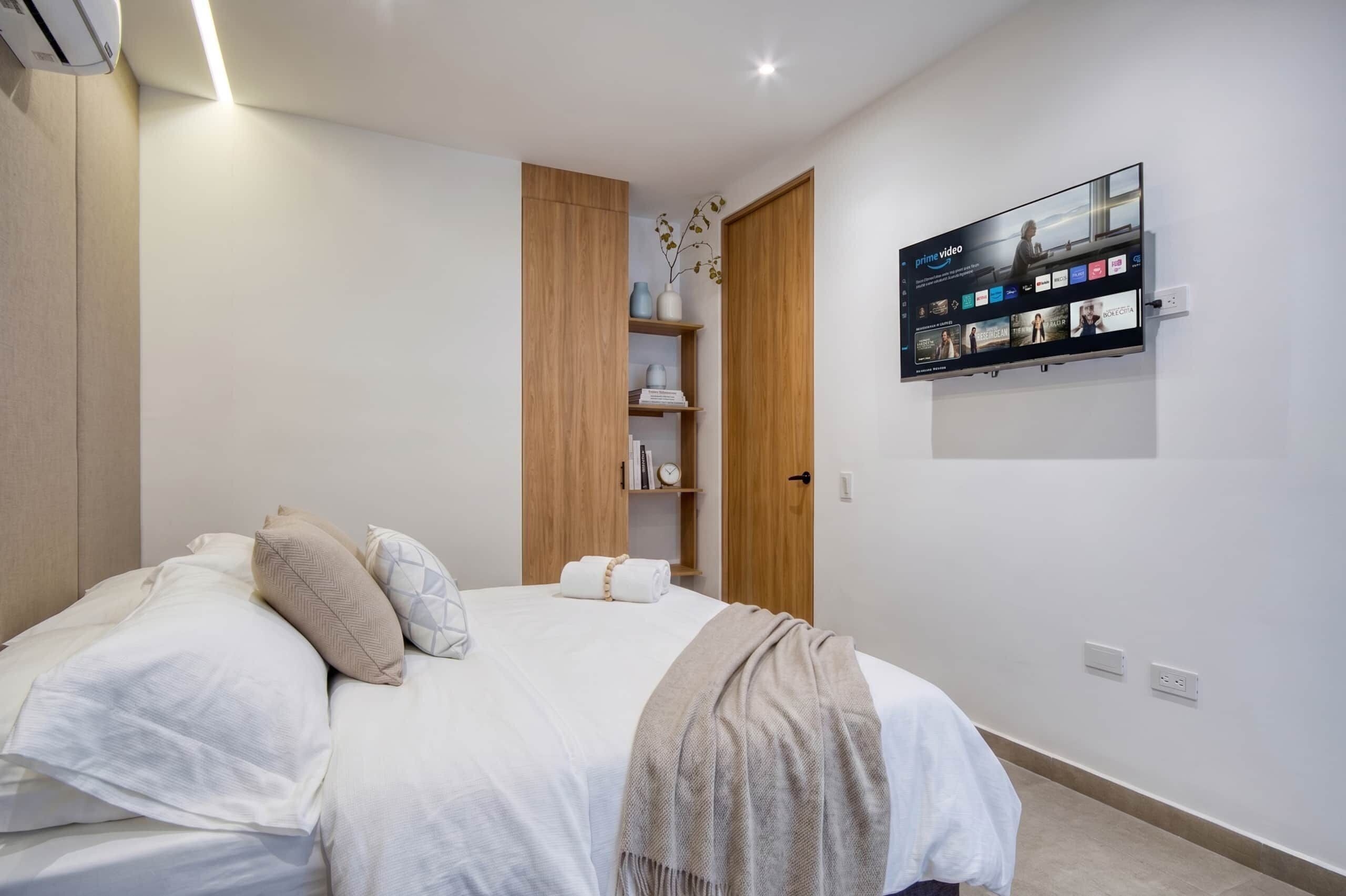 Quarto com cama, almofadas, mesa de cabeceira, prateleira com livros, televisão na parede e guarda-roupa de madeira.
