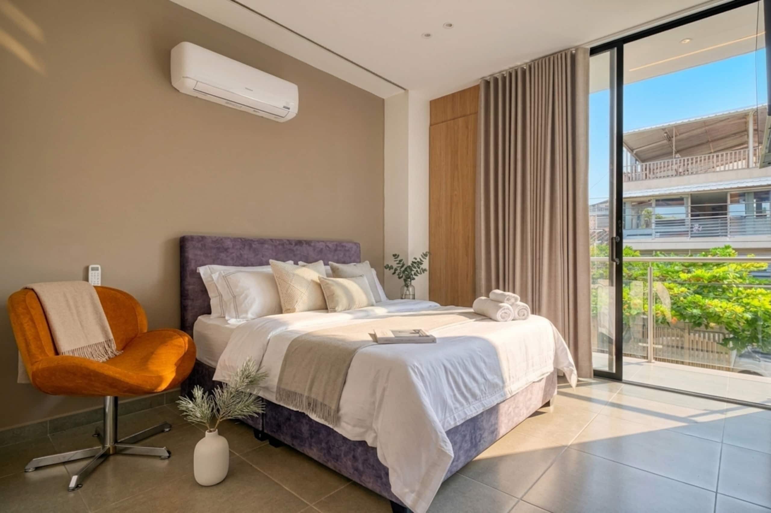 A modern bedroom with a bed, a chair, a potted plant, and a view of a building through a glass door.