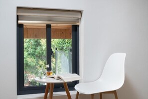 Quarto com uma grande janela, uma cadeira branca, uma pequena mesa com um livro e um copo de bebida, e vista para o verde lá fora.