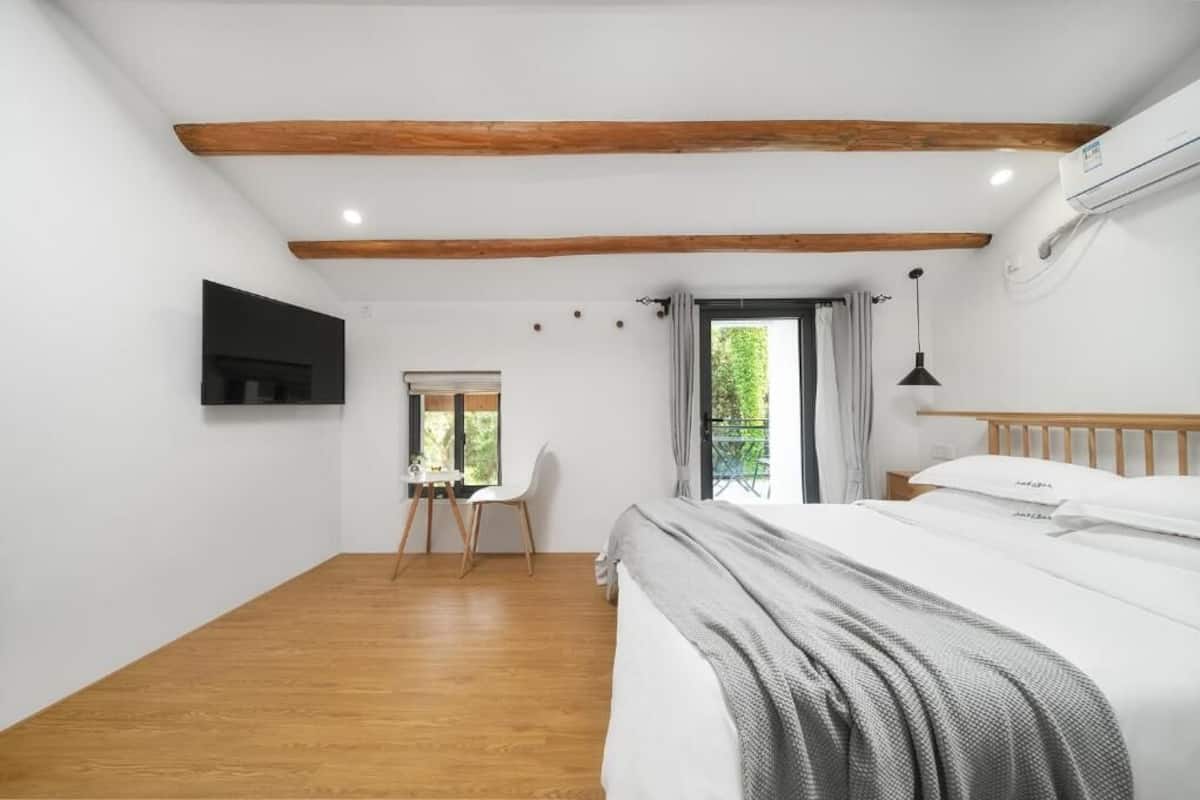 Quarto com cama, escrivaninha, cadeira, TV fixada na parede e janela com vista para o verde.