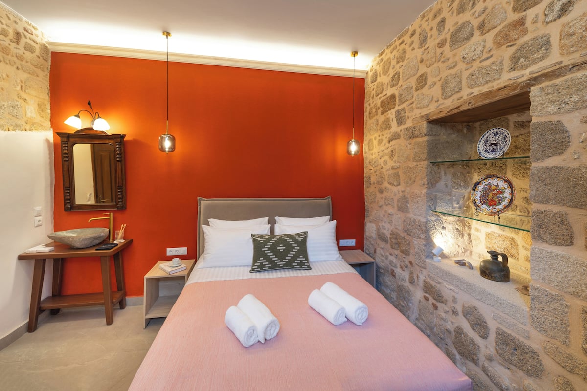 Quarto de hotel com parede de pedra, cama com roupa de cama branca, mesa de cabeceira, espelho e prateleira com pratos decorativos.