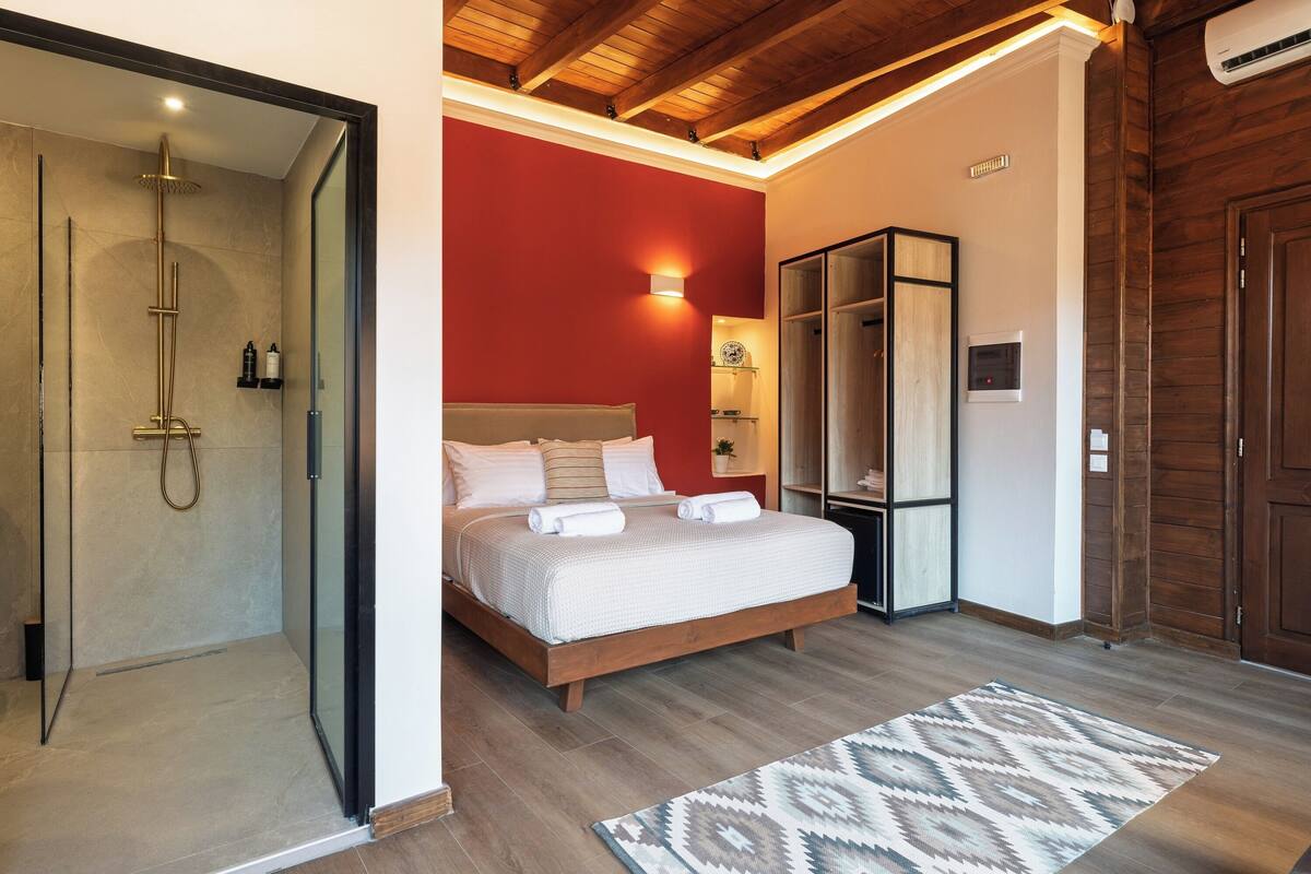 Quarto com cama, chuveiro, piso de madeira, tapete estampado e parede com detalhes em vermelho.