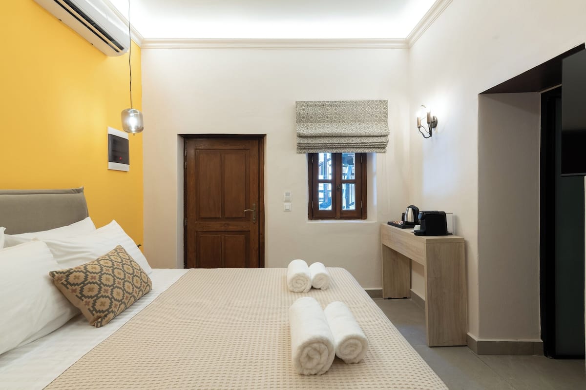Quarto de hotel com cama, escrivaninha, cafeteira e janela com persiana estampada.