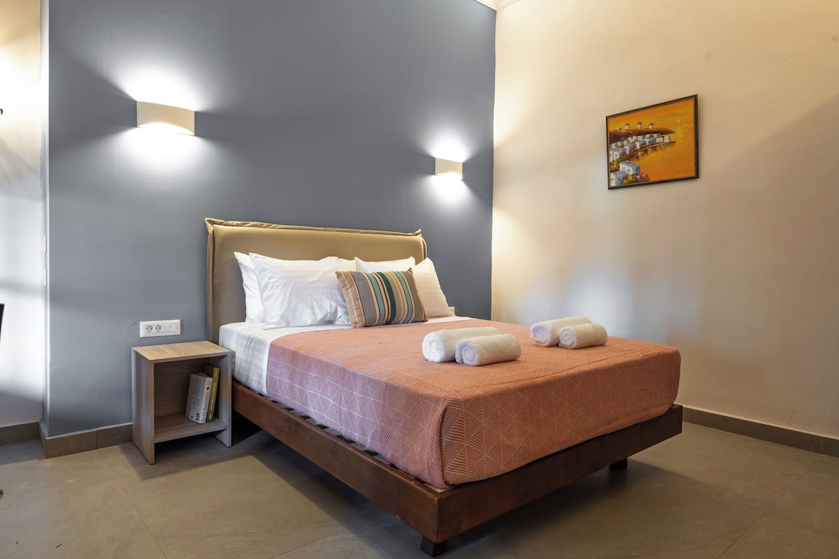 Cama bem arrumada com travesseiros e cobertor, mesa de cabeceira com um livro e luminária na parede.