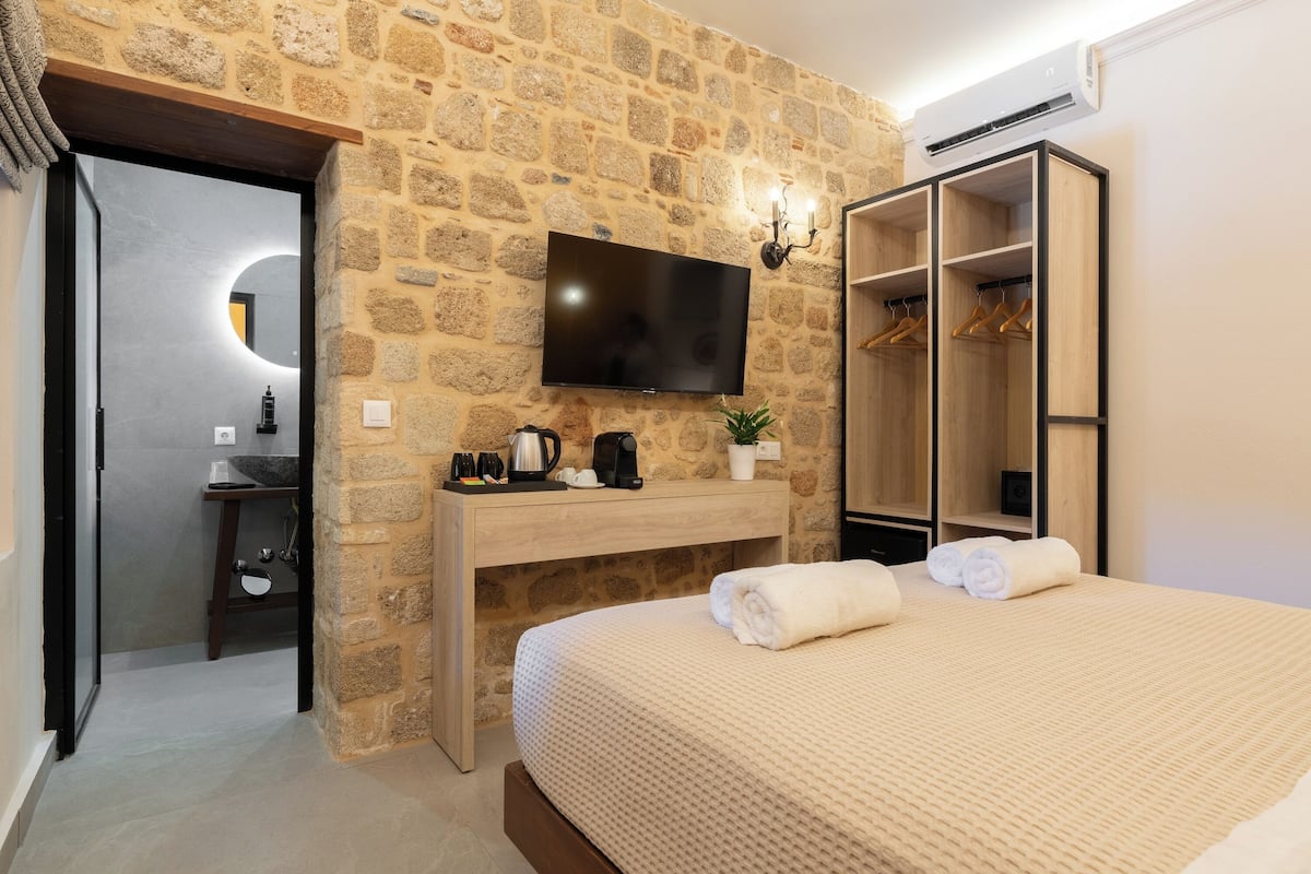 Quarto com parede de pedra, televisão de tela plana, guarda-roupa de madeira, cama com manta bege e uma pequena escrivaninha com cafeteira.