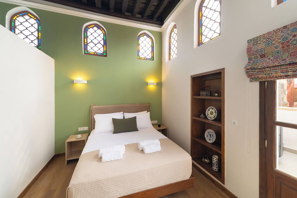 Quarto com cama, duas mesas de cabeceira, um criado-mudo, prateleira com pratos decorativos e três janelas ogivais com vitrais.