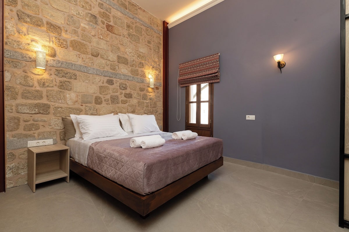 Um quarto com parede de pedra, uma cama com colchão acolchoado, duas luminárias de cabeceira, uma janela com persiana e uma mesinha de cabeceira de madeira.