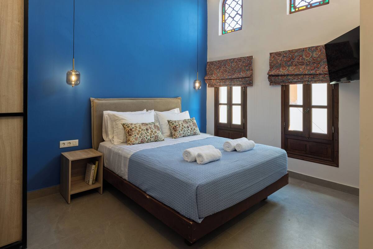 Quarto com parede azul, estrutura de cama de madeira, mesa de cabeceira com um livro e janela com cortina estampada.