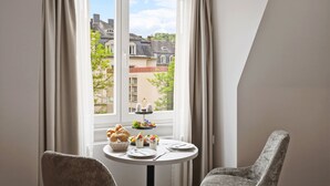 Quarto com mesa de jantar posta para dois, janela com cortinas e vista para prédios.
