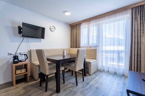Quarto com televisão na parede, mesa lateral de madeira com cafeteira, mesa de jantar com cadeiras e um sofá.