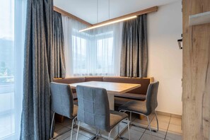 Uma área de refeições moderna com mesa de madeira e cadeiras estofadas em cinza, janelas amplas com cortinas leves e vista para o exterior.