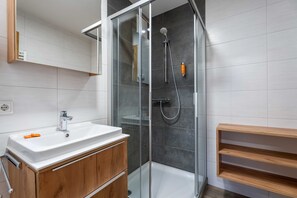 Uma casa de banho moderna com chuveiro envidraçado, lavatório branco, armários de madeira e prateleira embutida.