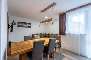Uma sala de jantar moderna com uma mesa de madeira, cadeiras pretas, um lustre no teto, um relógio fixado na parede e uma janela com cortinas leves.