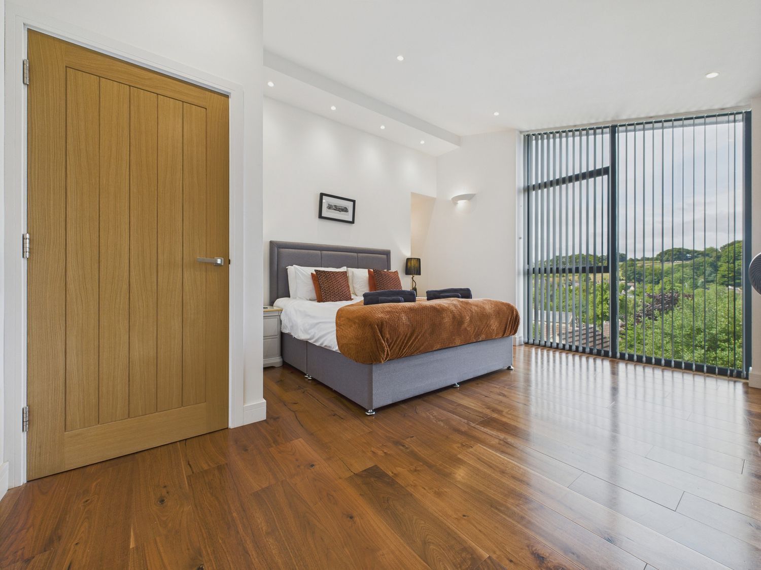 Quarto com porta de madeira, cama com cabeceira cinza, mesa de cabeceira, luminária e vista através de uma janela grande.
