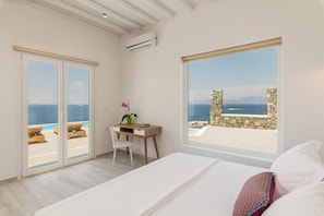 Quarto com cama, escrivaninha, cadeira e grandes janelas com vista para o mar e uma parede de pedra.