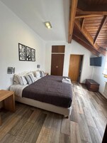 Quarto com cama, piso de madeira, ventoinha no teto, televisão e porta.