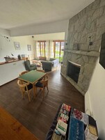 Uma sala de estar com lareira de pedra, área de jantar com mesa e cadeiras verdes, e vista para o exterior por grandes janelas.