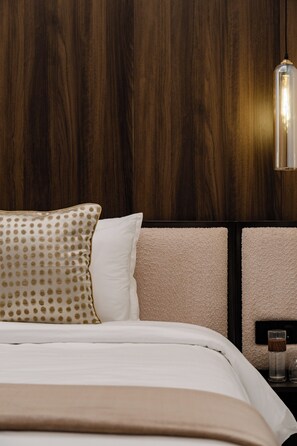 Cama bem arrumada com roupa de cama branca e bege, cabeceira de madeira e luminária fixada na parede.