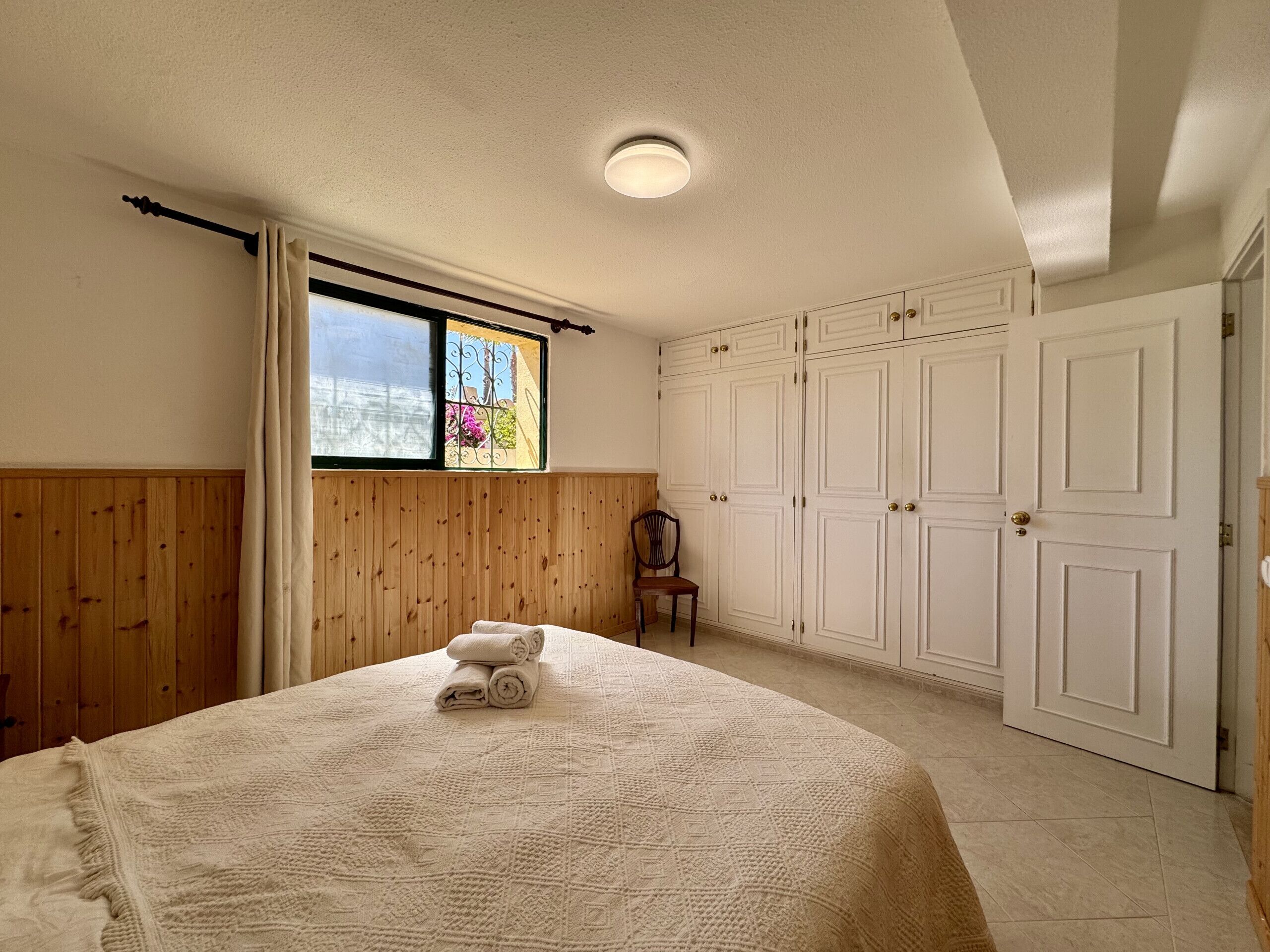 Um quarto com cama, janela com cortinas, uma cadeira e um guarda-roupa.