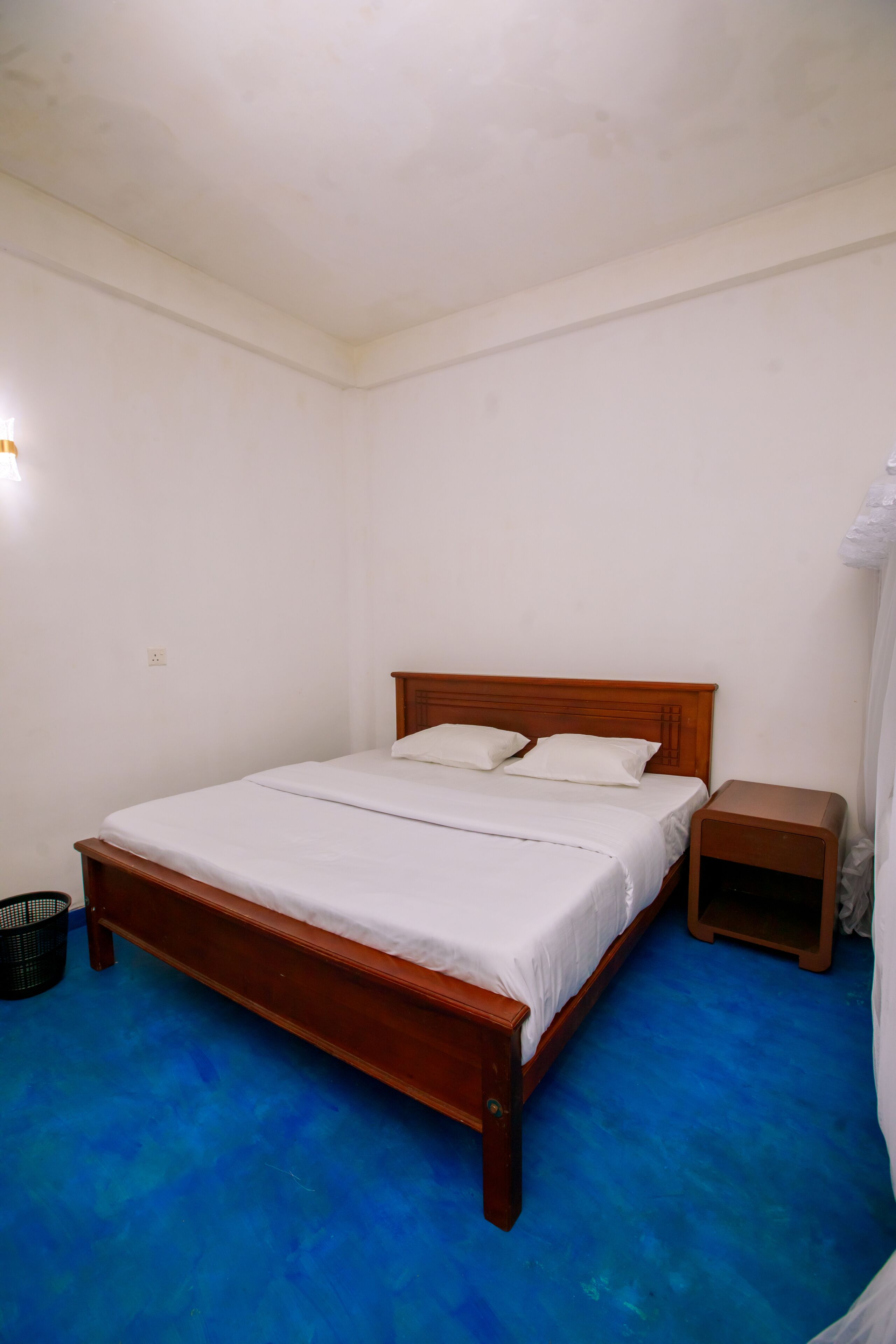 Uma cama de solteiro com cabeceira de madeira, mesa de cabeceiro e chão coberto por um tapete azul em um quarto com paredes brancas.