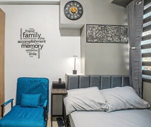 Quarto de hotel com cama, uma cadeira azul, mesa de cabeceira com abajur, relógio de parede e uma obra de arte com nuvem de palavras.