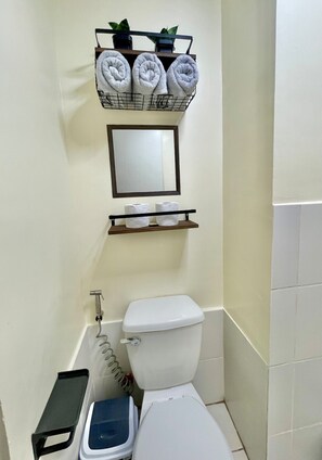 Um banheiro com vaso sanitário, uma prateleira com toalhas, um espelho pequeno e uma planta.