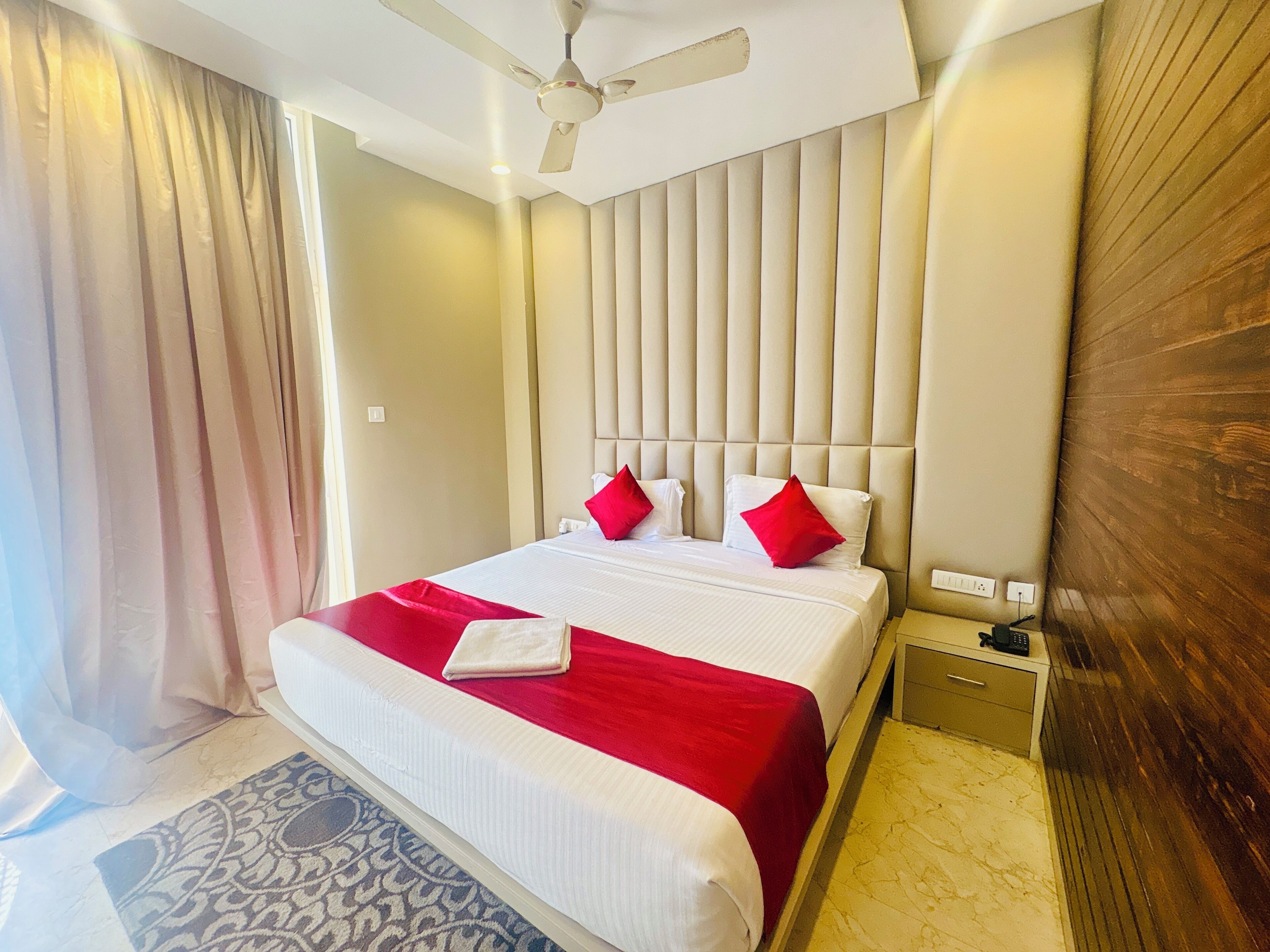 Quarto de hotel com cama, mesa de cabeceira, ventoinha no teto, janela com cortinas e tapete estampado.