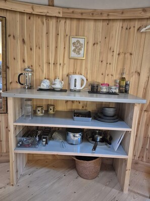 Interior em madeira com prateleiras contendo utensílios de cozinha, uma cafeteira e um quadro emoldurado.