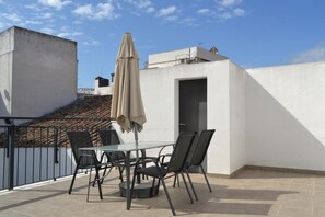 Um terraço no telhado com mesa e cadeiras de vidro, um guarda-sol bege e uma parede branca.