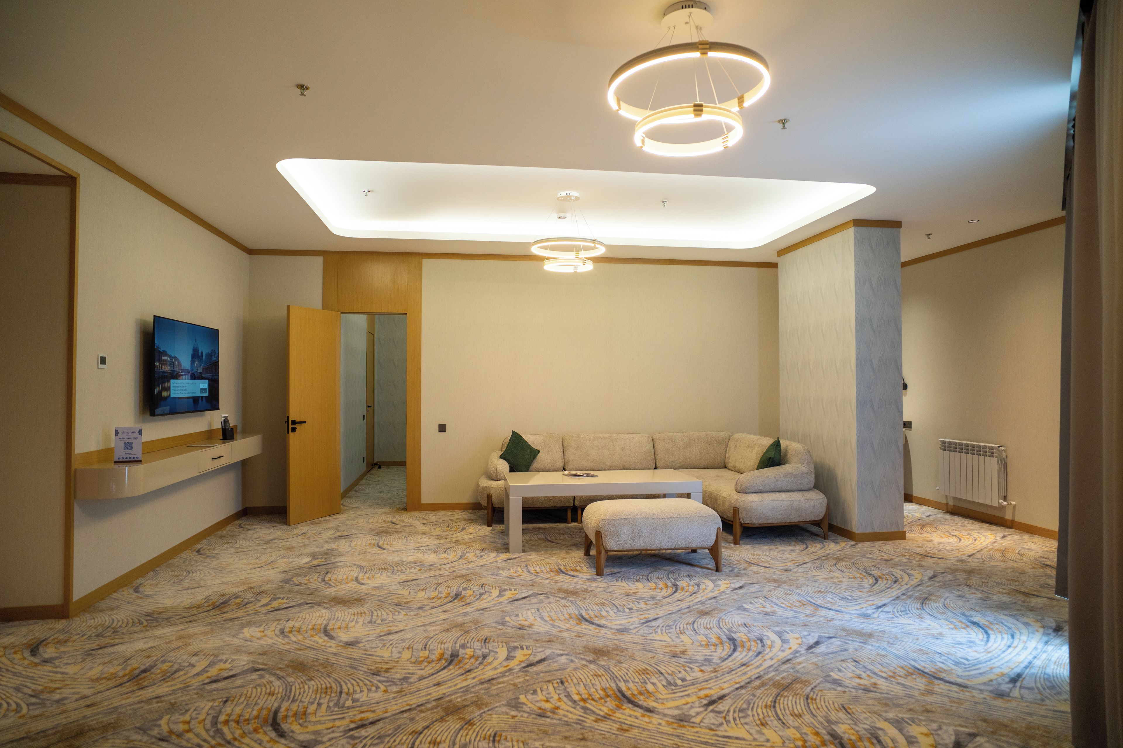 Quarto de hotel moderno com sofá, banqueta e TV de tela plana sobre um console de madeira.