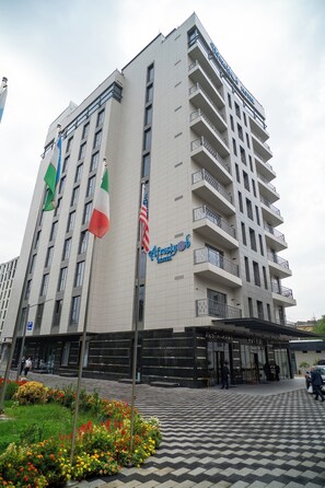 Um hotel com vários andares, um mastro com bandeiras e um pavimento quadriculado em frente.