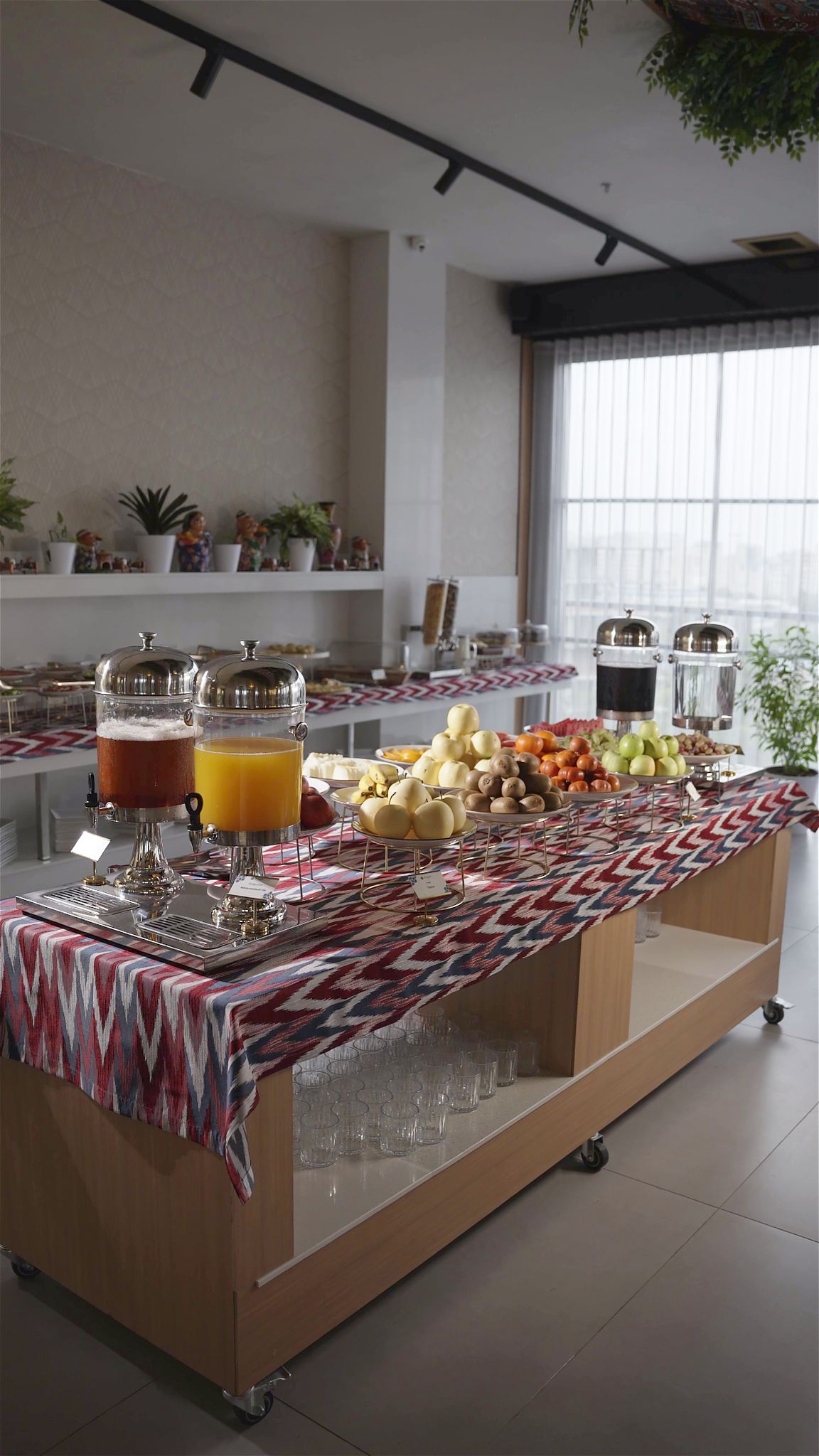 Disposição de buffet com estação de café, frutas e toalha de mesa estampada.