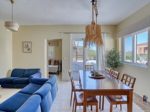 Uma sala de estar moderna com um sofá azul, uma mesa de jantar de madeira com cadeiras, um lustre e vista para o exterior.