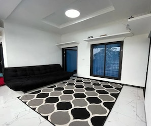 Uma sala de estar com um sofá preto, um tapete estampado, teto branco e uma janela com vista.