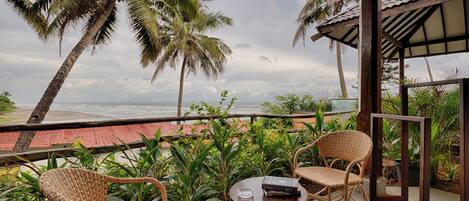 Uma área de estar coberta ao ar livre, com cadeiras de vime e uma mesa, com vista para uma praia com palmeiras e o oceano.